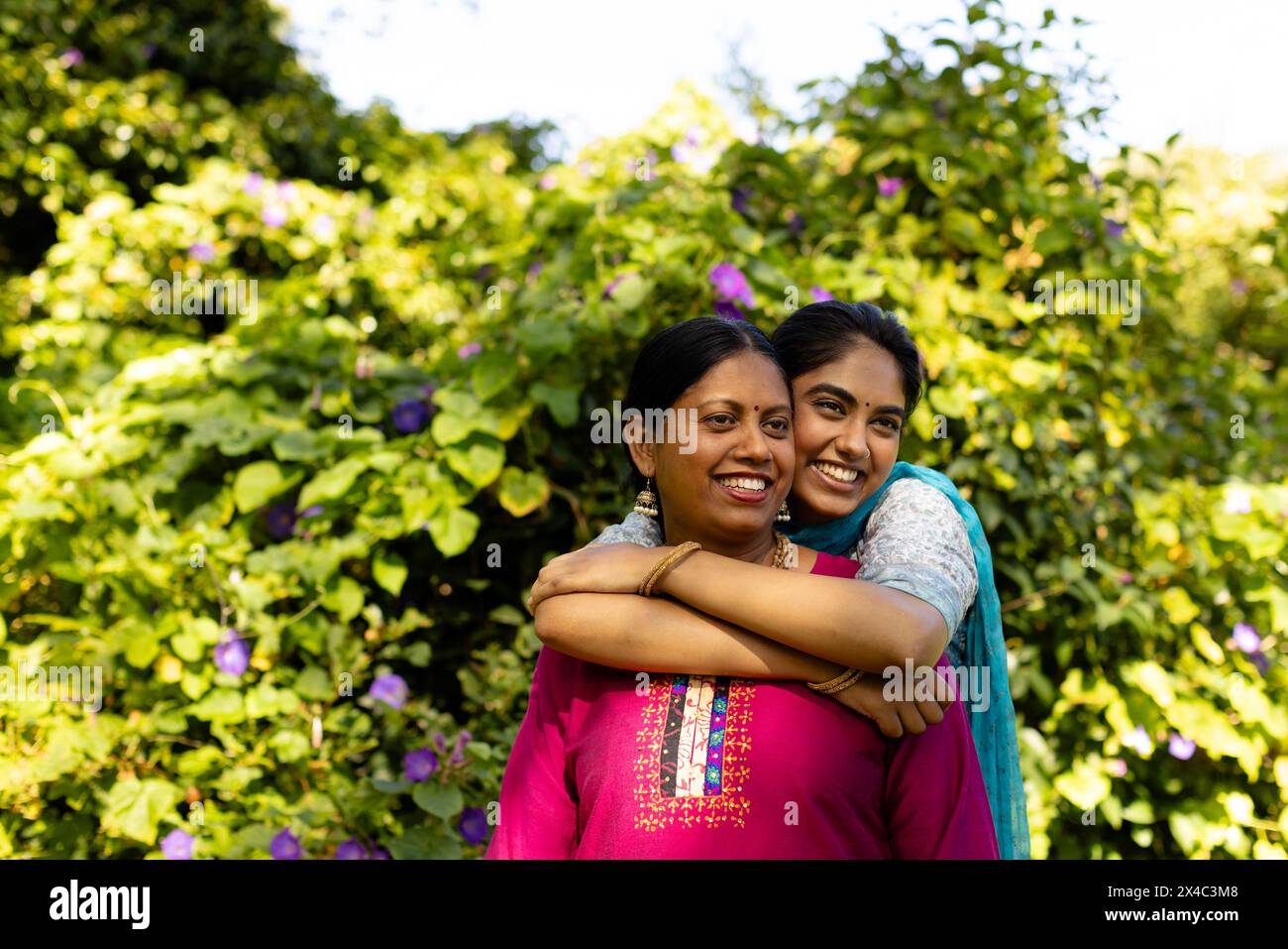 Indian mother and teenage daughter hugging, both smiling in garden, copy space. Mother with ...