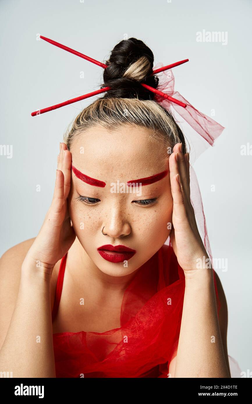 A striking Asian woman adorned in red makeup and vibrant attire, dramatically holds her head ...