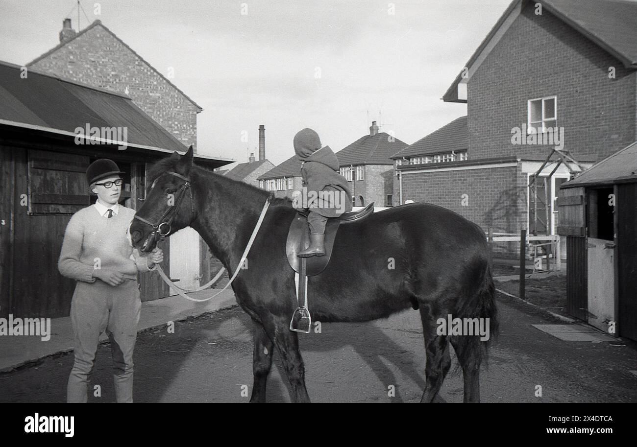 1960s, historical, outside at a suburban riding school, an infant child ...