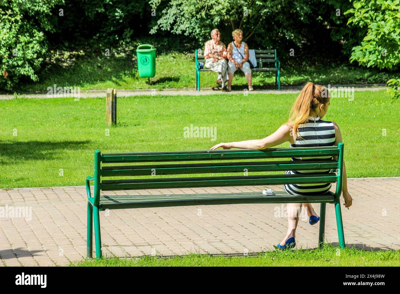 Young woman sitting on the bench opposite elderly couple in public park ...