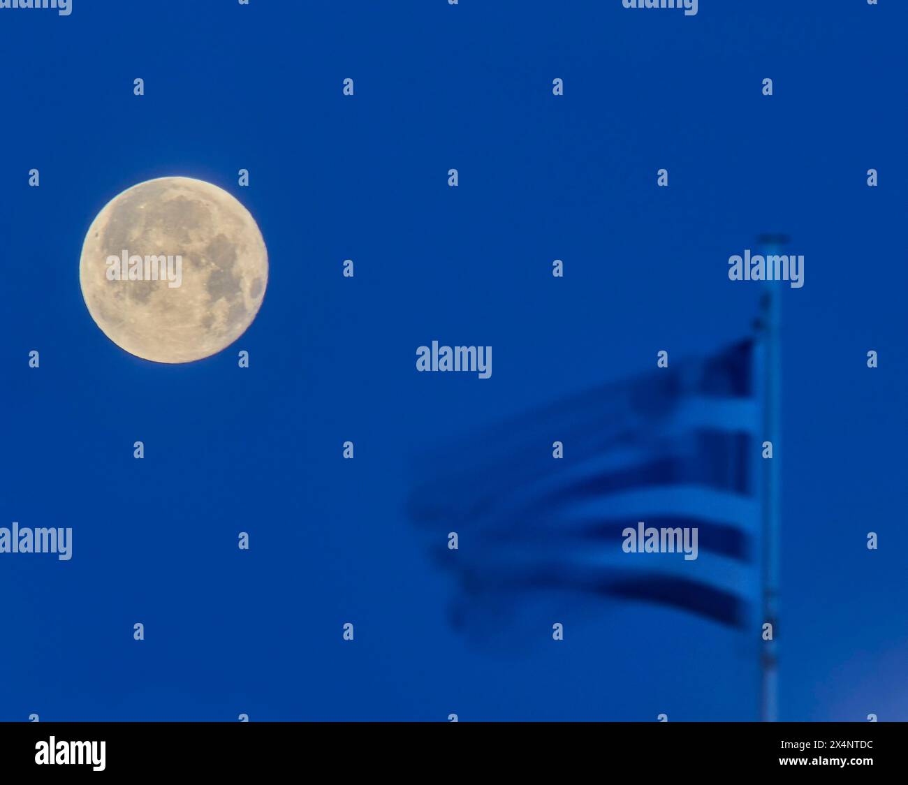 A full moon next to a waving Greek flag in the blue hour, twilight ...