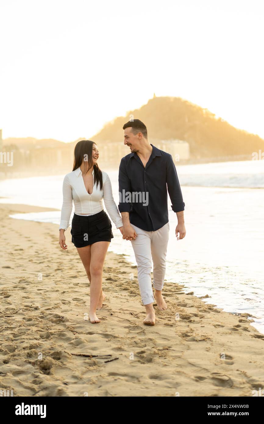 Vertical photo of a young caucasian chic couple walking barefoot holding hands along a beach ...