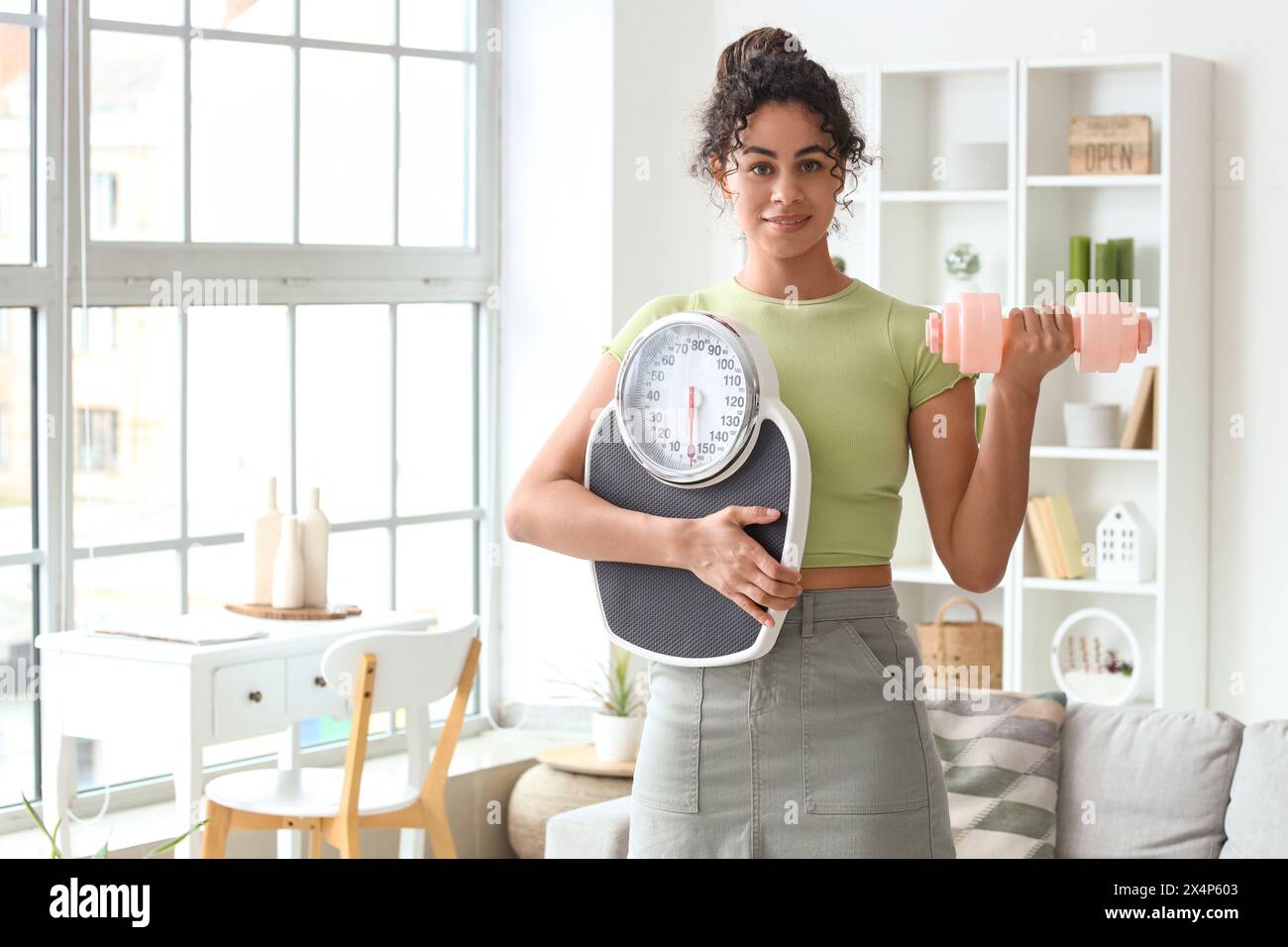 Young African-American woman with weight scales and dumbbell at home ...