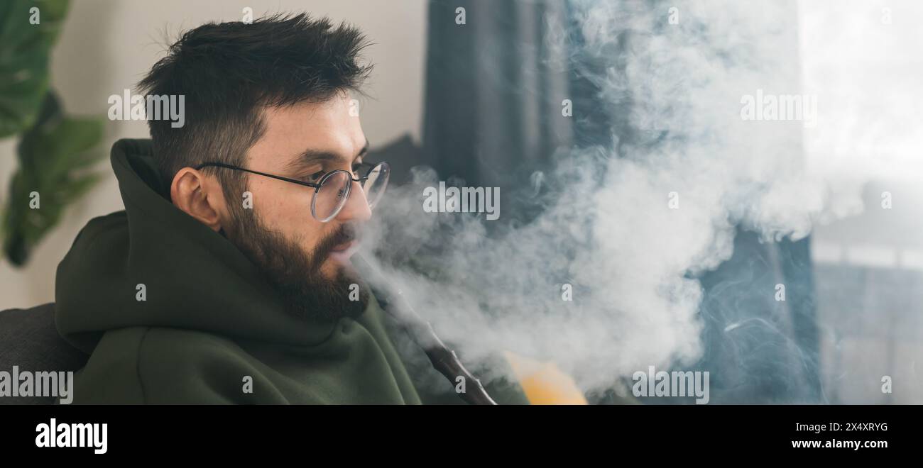 Bearded millennial or gen z man smoking hookah while relaxing on sofa ...