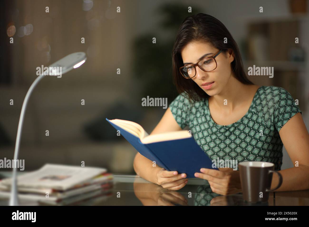 Happy woman wearing eyeglass reading a paper book in the night at home ...