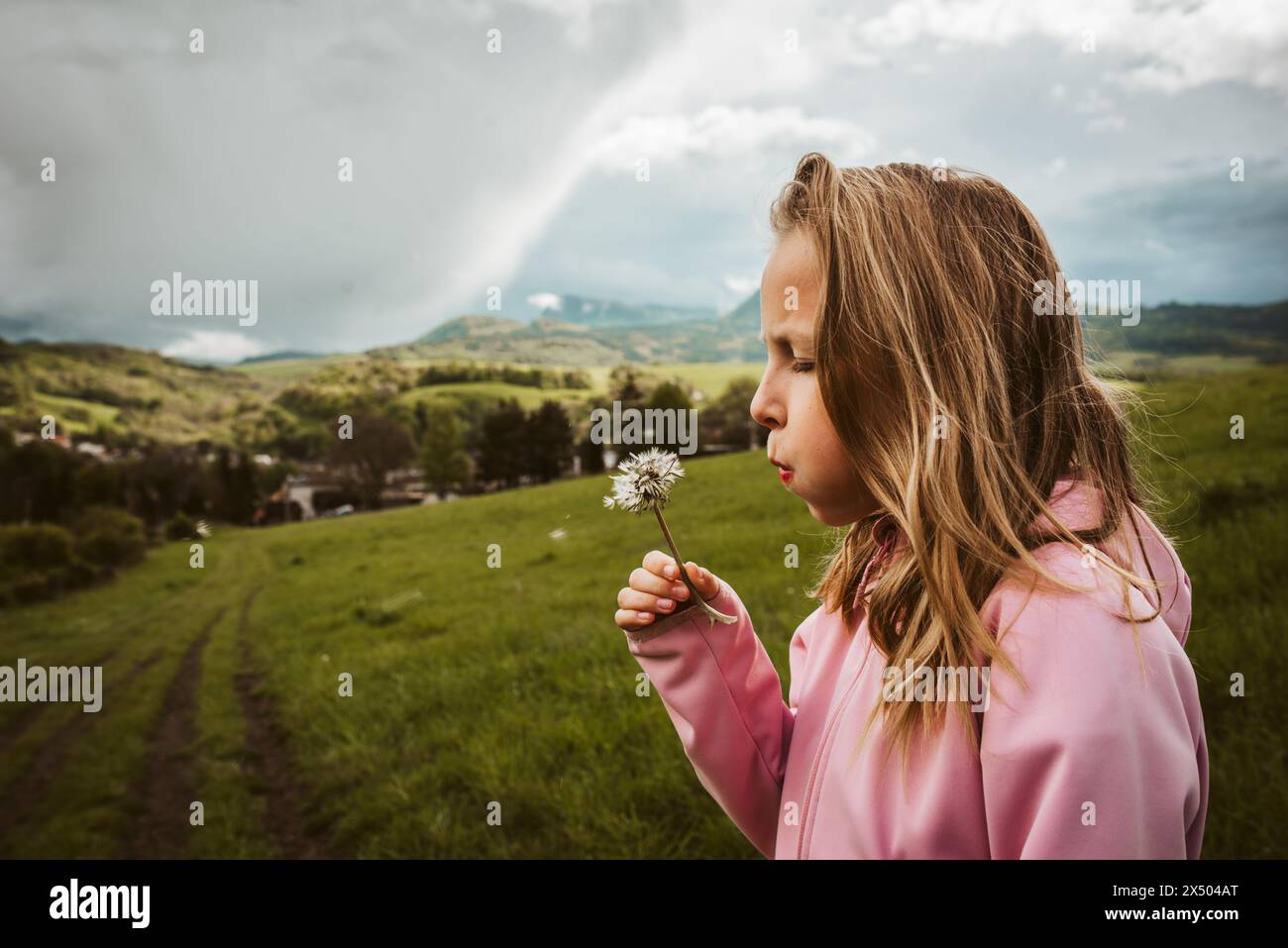 Girl blowing a dandelion in spring nature, rain clouds on sky. Wearing ...