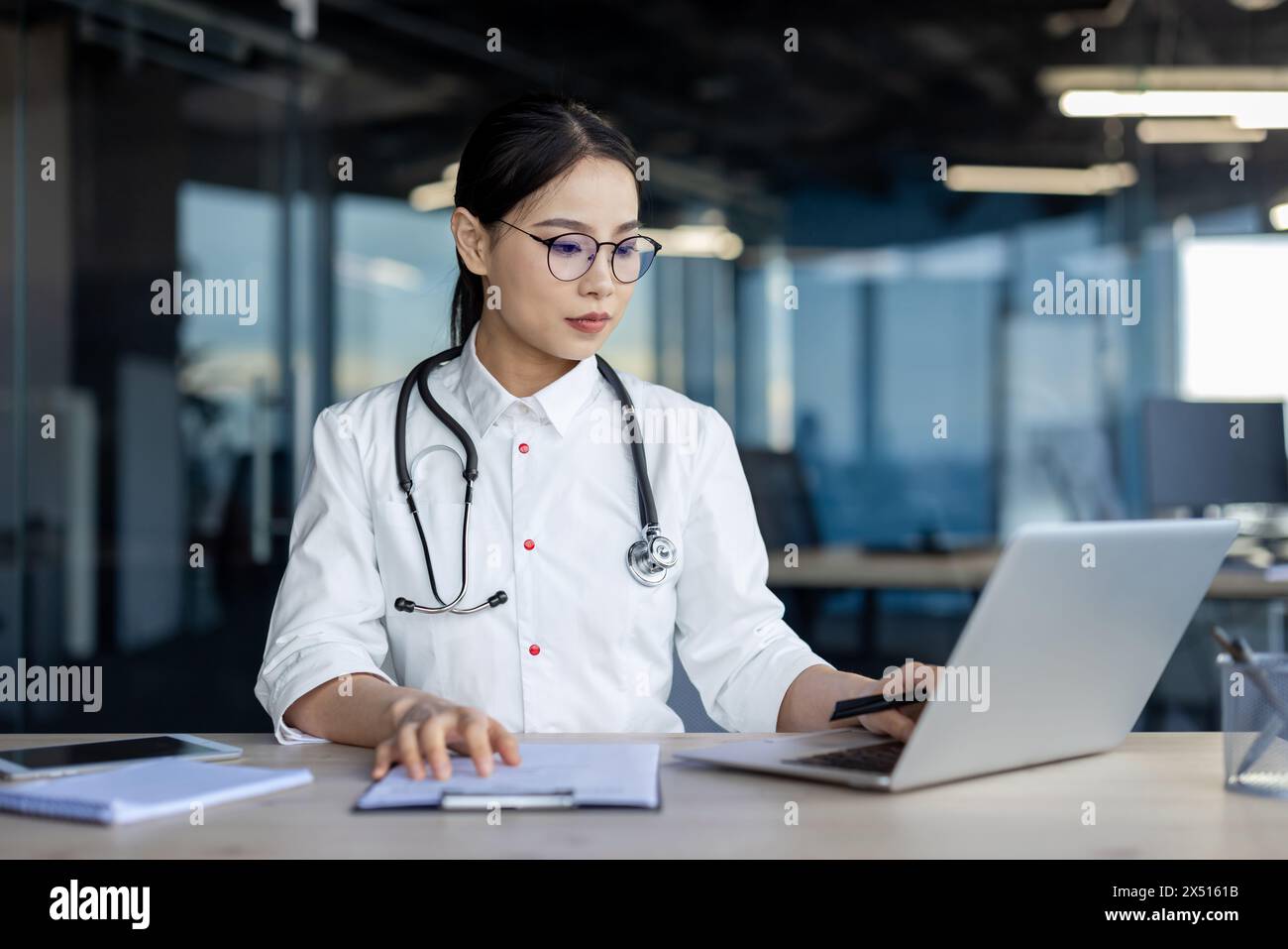 A focused Asian female doctor with glasses analyzes data on her laptop ...