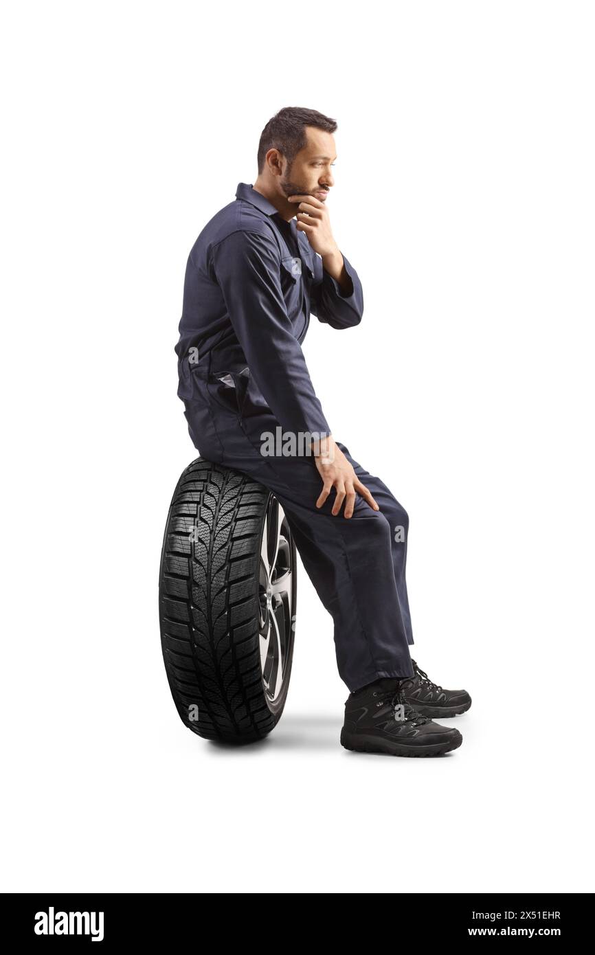 Pensive car mechanic in a uniform sitting on a tire isolated on white ...