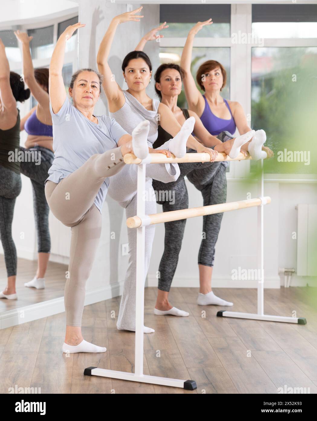Senior woman performing stretching exercises at ballet barre with group ...