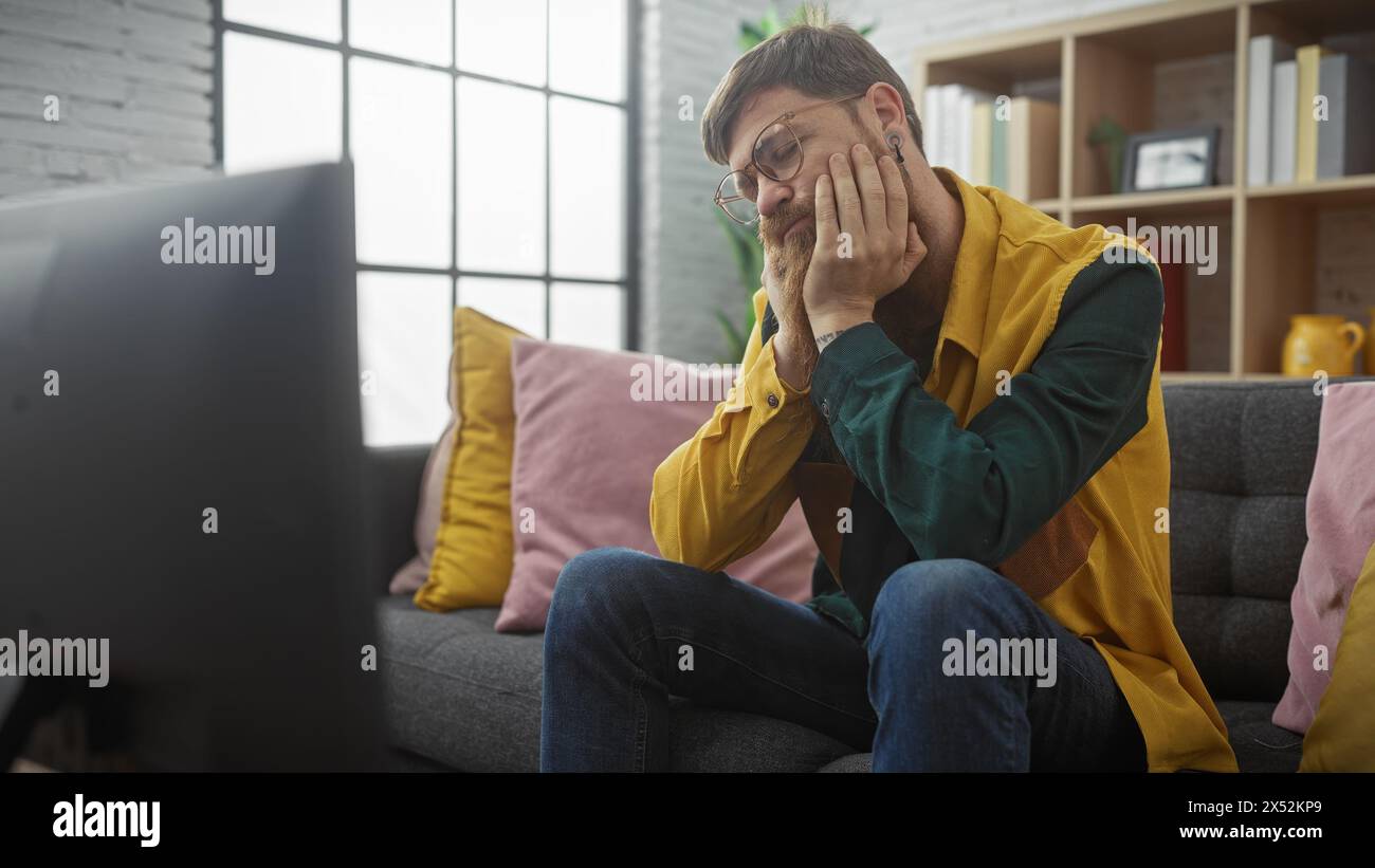 A bored young man with glasses and a beard sitting on a grey sofa ...