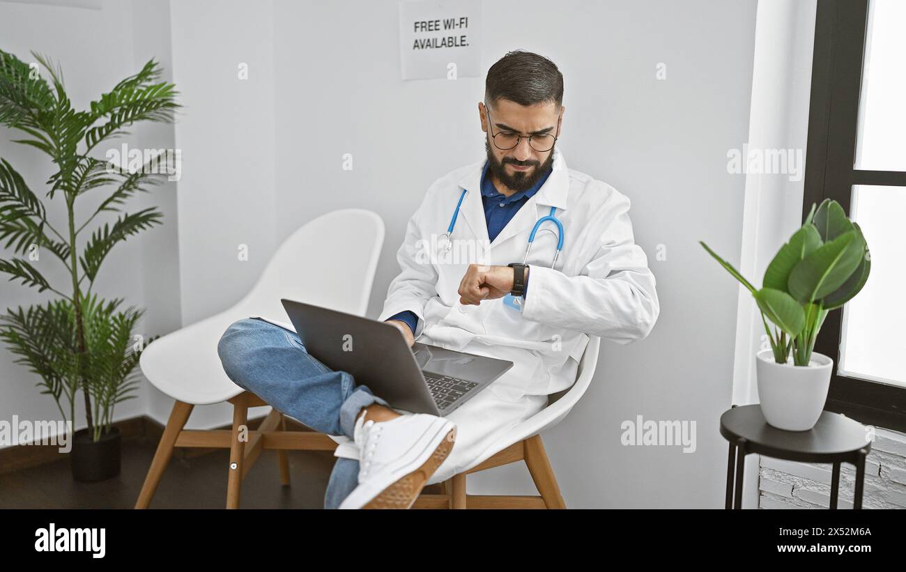 Bearded man in lab coat checks watch while working on laptop in modern ...