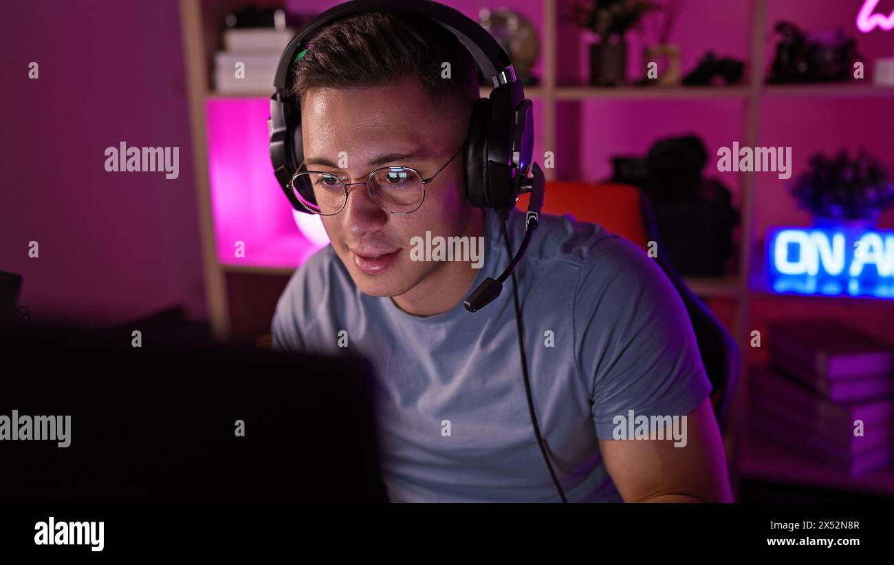 A focused hispanic man with headphones sits in a dark gaming room at ...