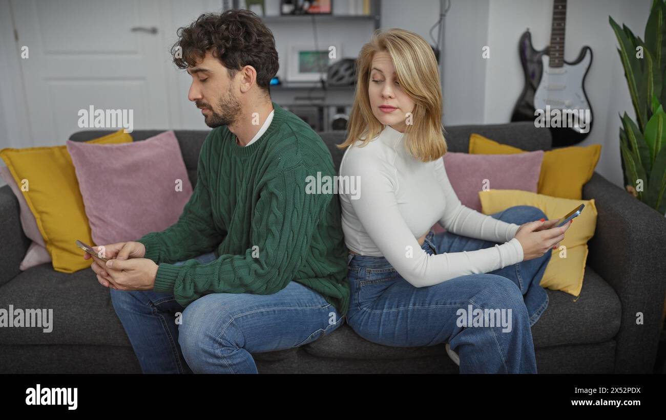 A woman sneakily glances at a man's phone while both sit back-to-back on a couch indoors with ...