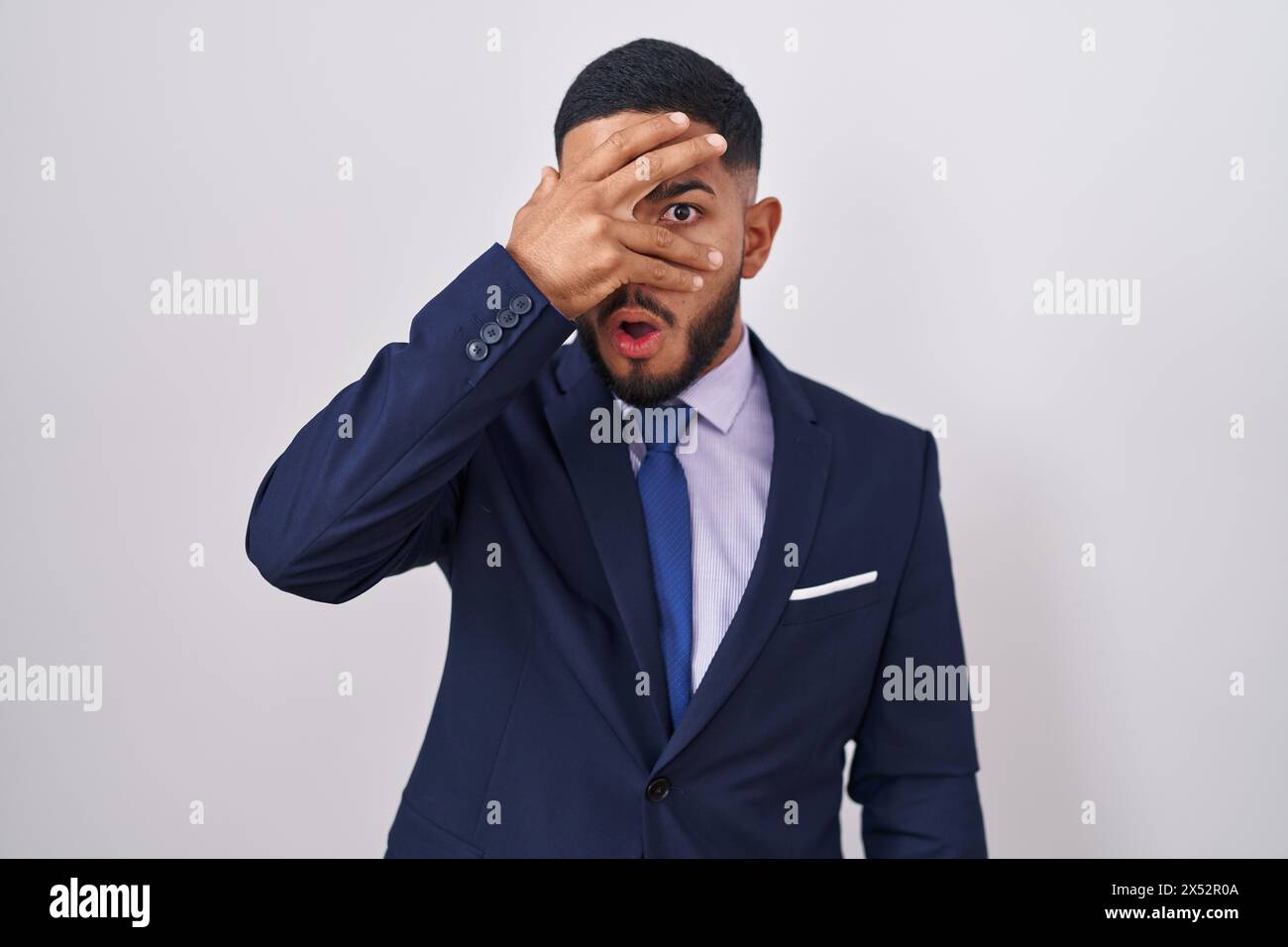 Young hispanic man wearing business suit and tie peeking in shock ...