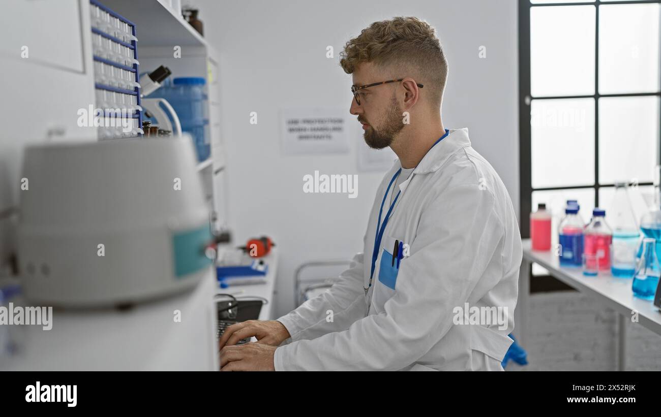 A young, handsome caucasian man with a beard and blue eyes wearing a lab coat works in a modern ...