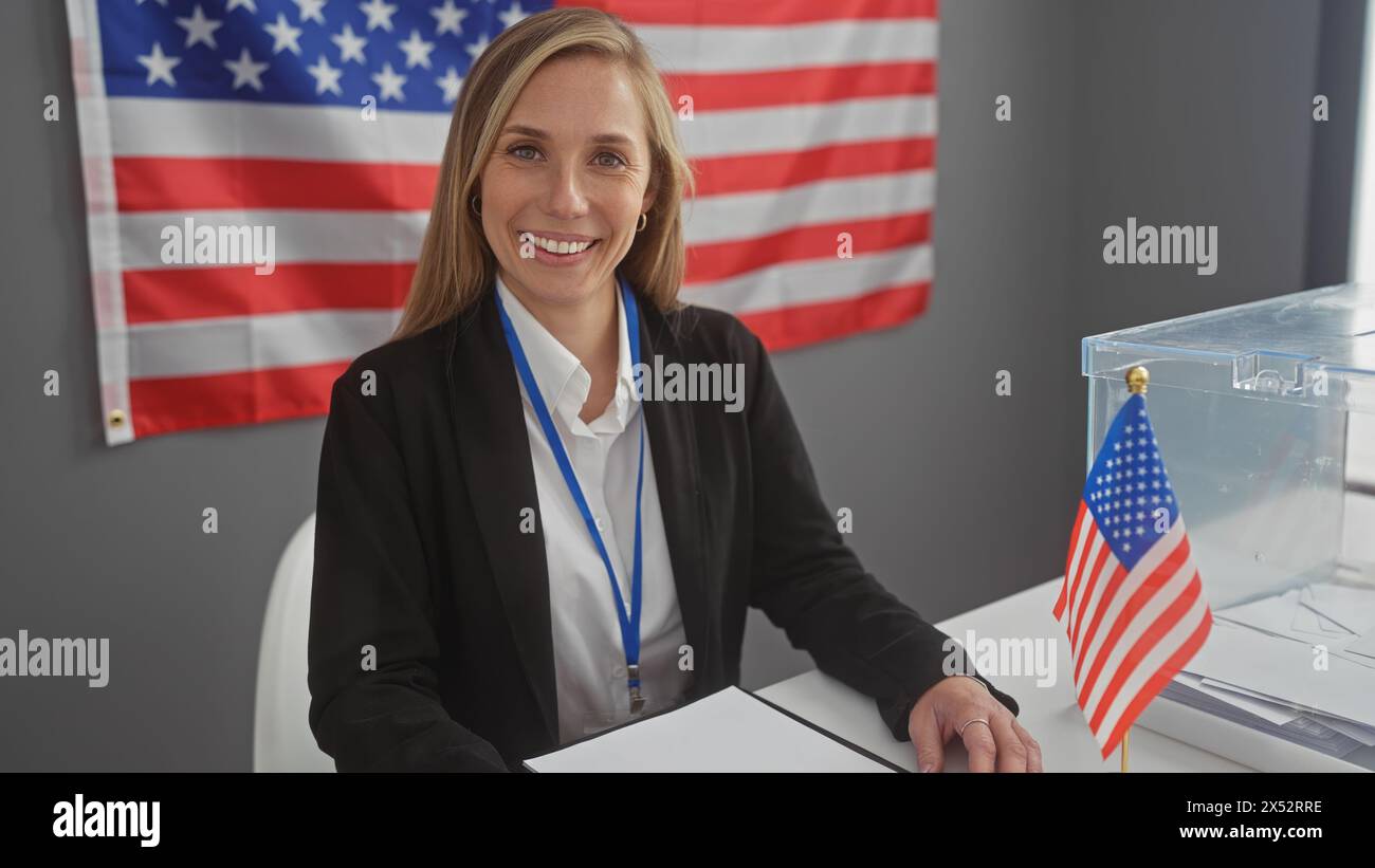 A smiling woman with a badge stands by a ballot box in a room with the ...