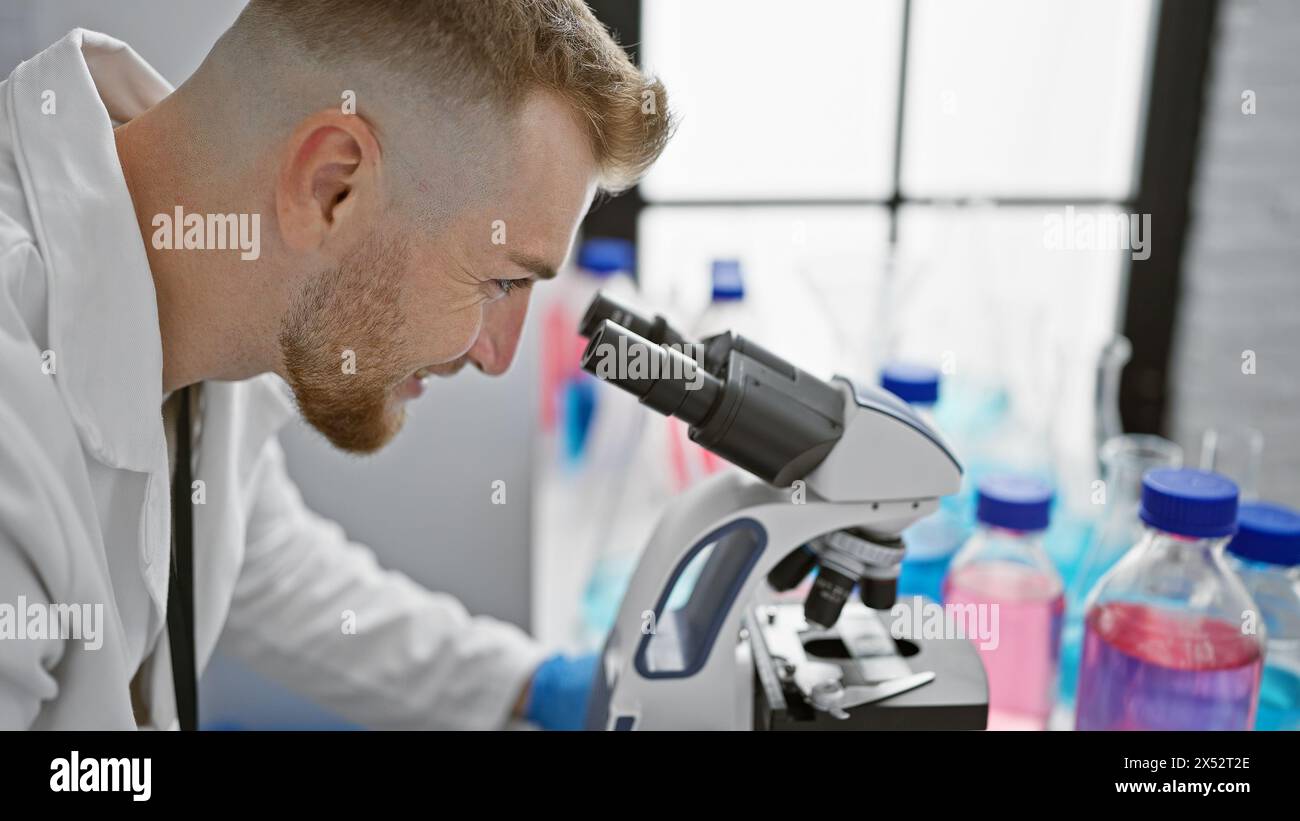 A concentrated young bearded man wearing a white lab coat working with a microscope in a science ...