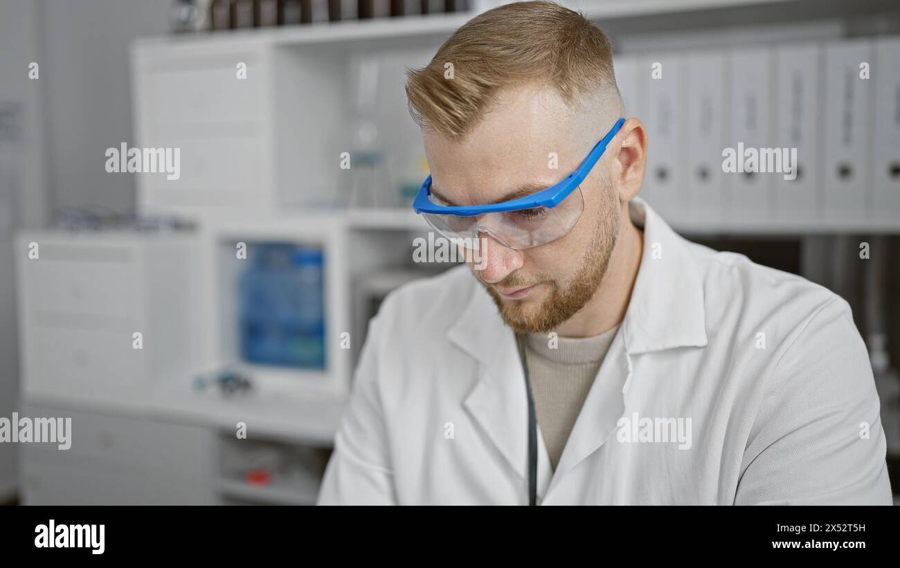 A focused young man with a beard wearing safety goggles and a lab coat ...