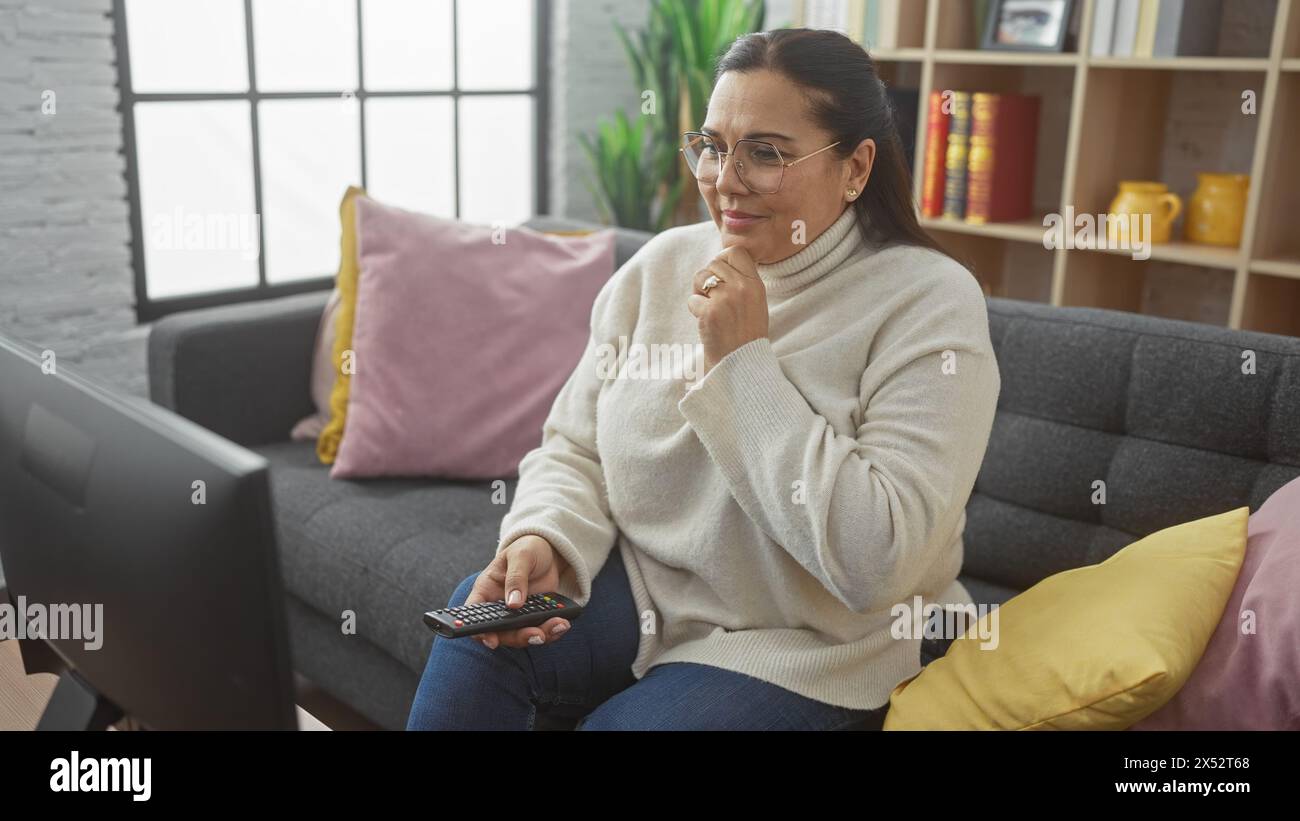 A smiling middle-aged hispanic woman relaxing in her modern living room holding a remote Stock ...