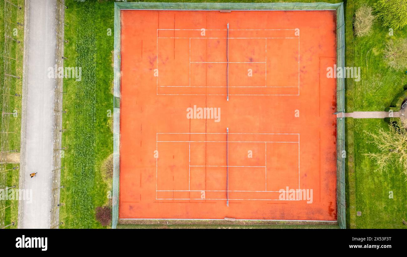 This aerial image features an unoccupied red clay tennis court ...