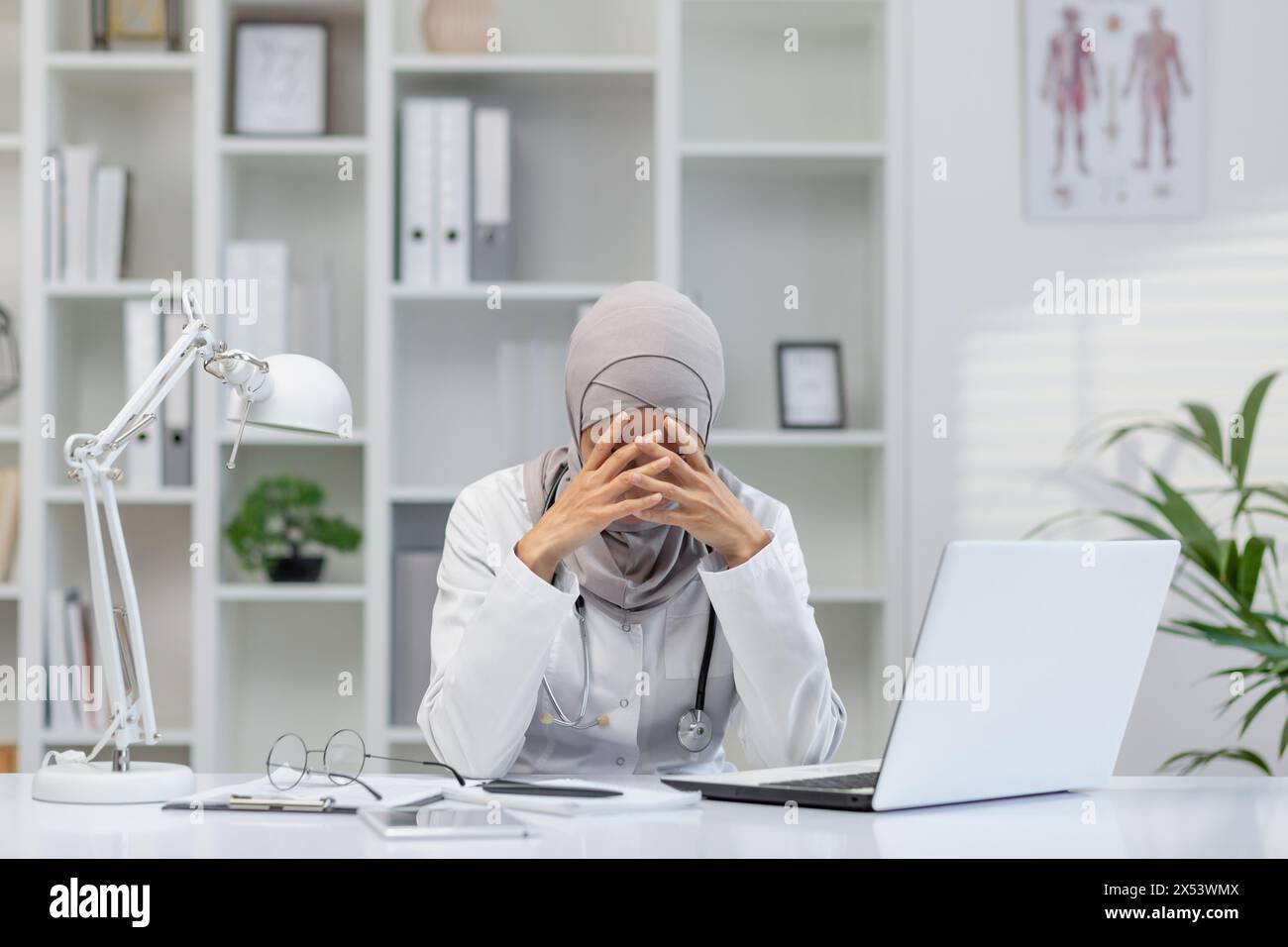 A female doctor in a hijab sits at her office desk, visibly stressed ...
