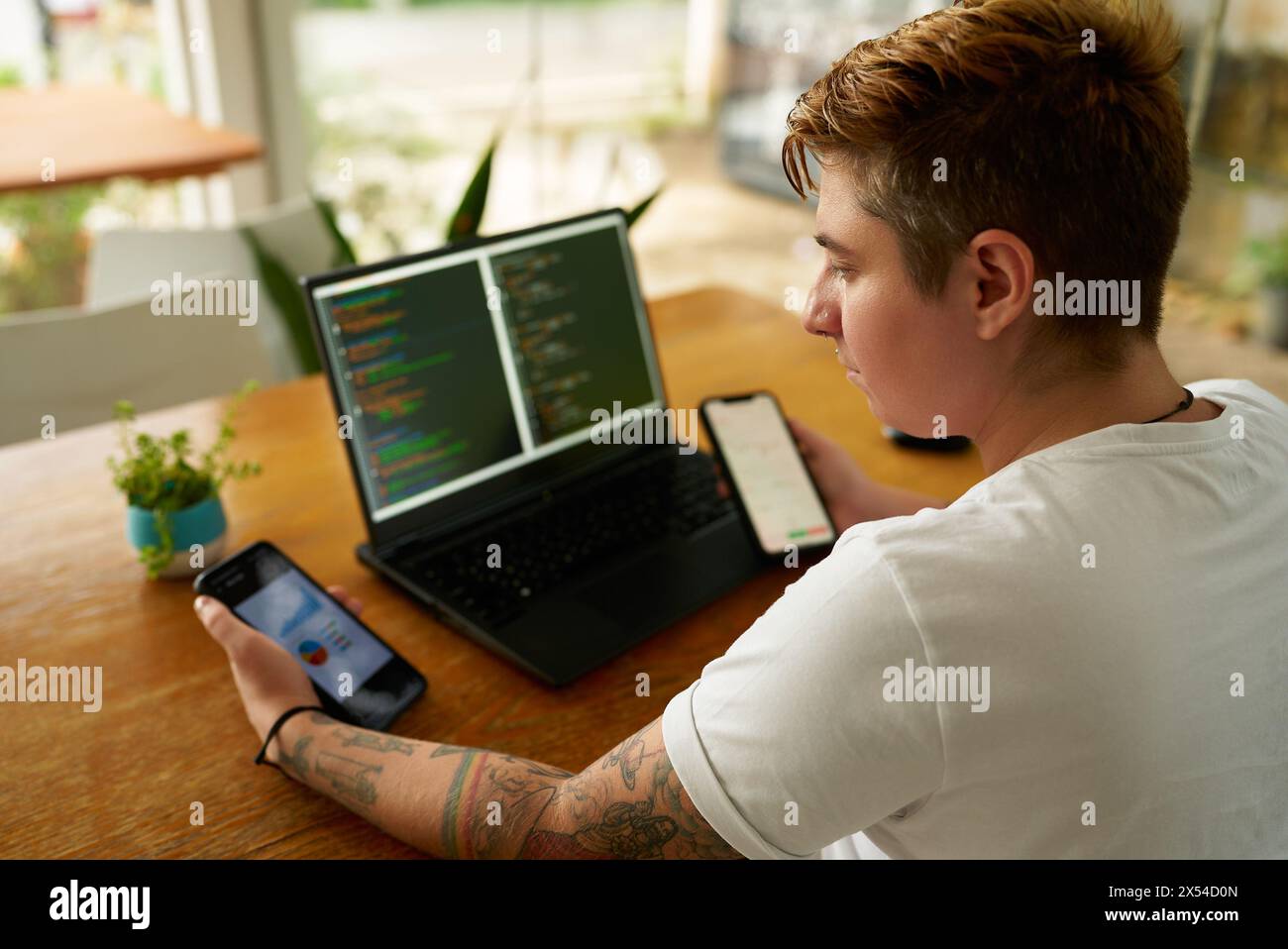 Person at wooden desk engages in intense multitasking with two smartphones displaying graphs ...