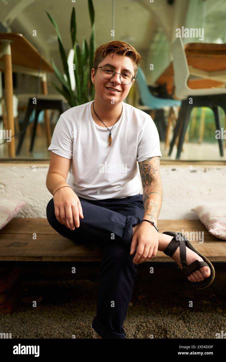 Transgender individual in smart casual attire sits smiling in modern ...