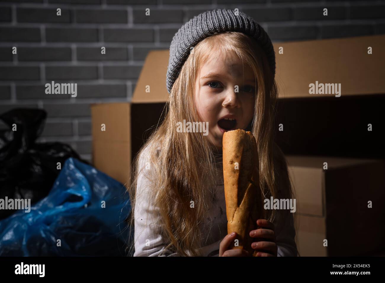 Homeless little girl eating bread near trash against grey brick wall ...