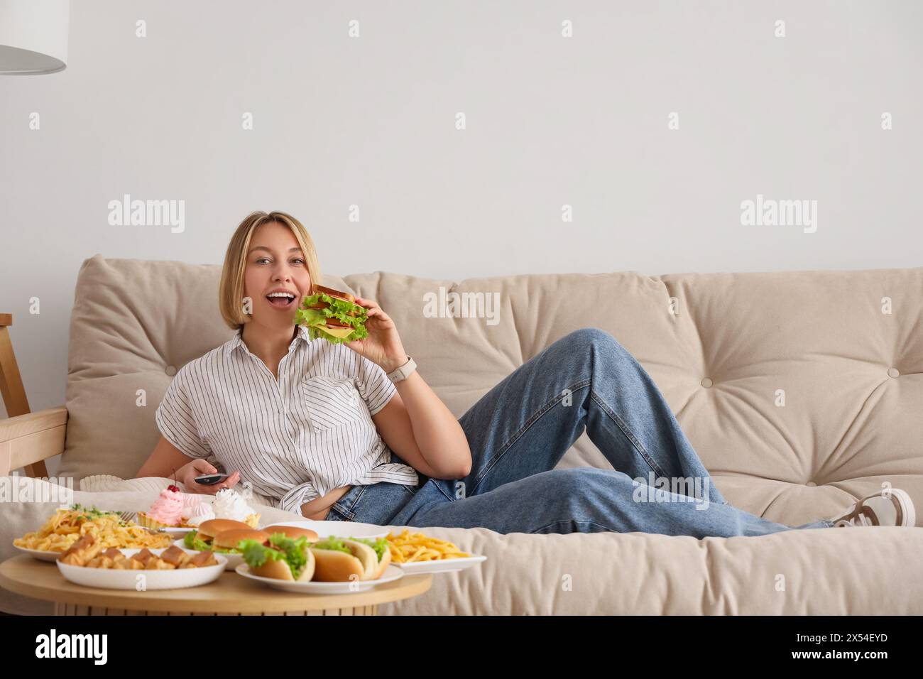 Beautiful woman with big sandwich watching TV on sofa at home ...