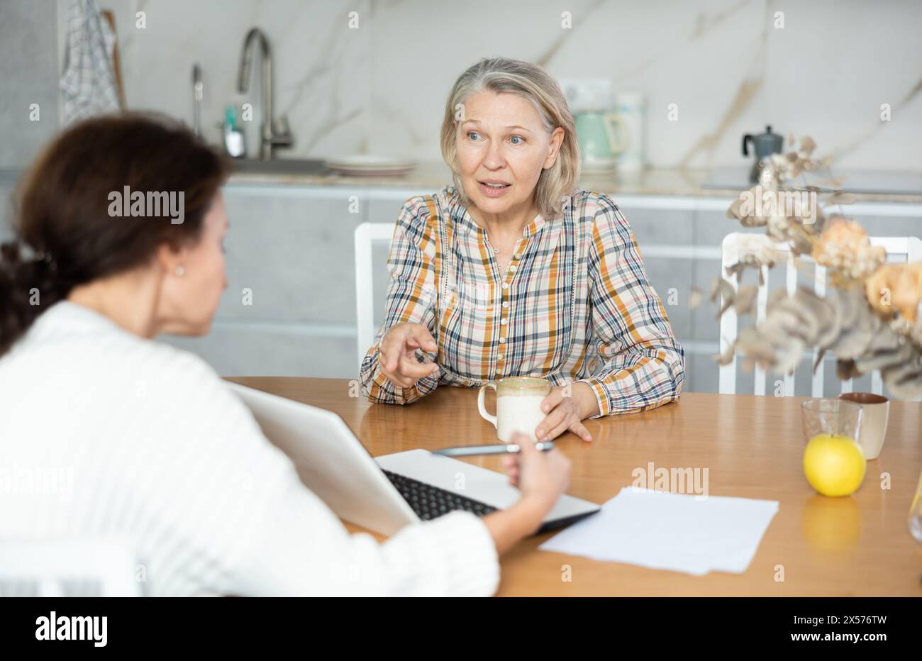 Positive old woman sitting at the kitchen-table face to face with ...