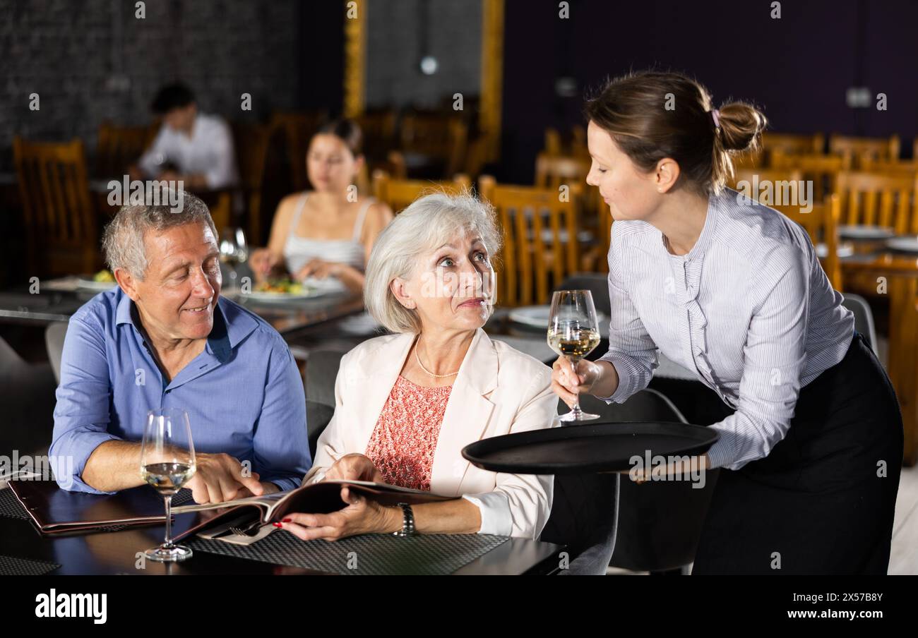 Waitress bringing aperitif to elderly couple selecting dishes from menu ...