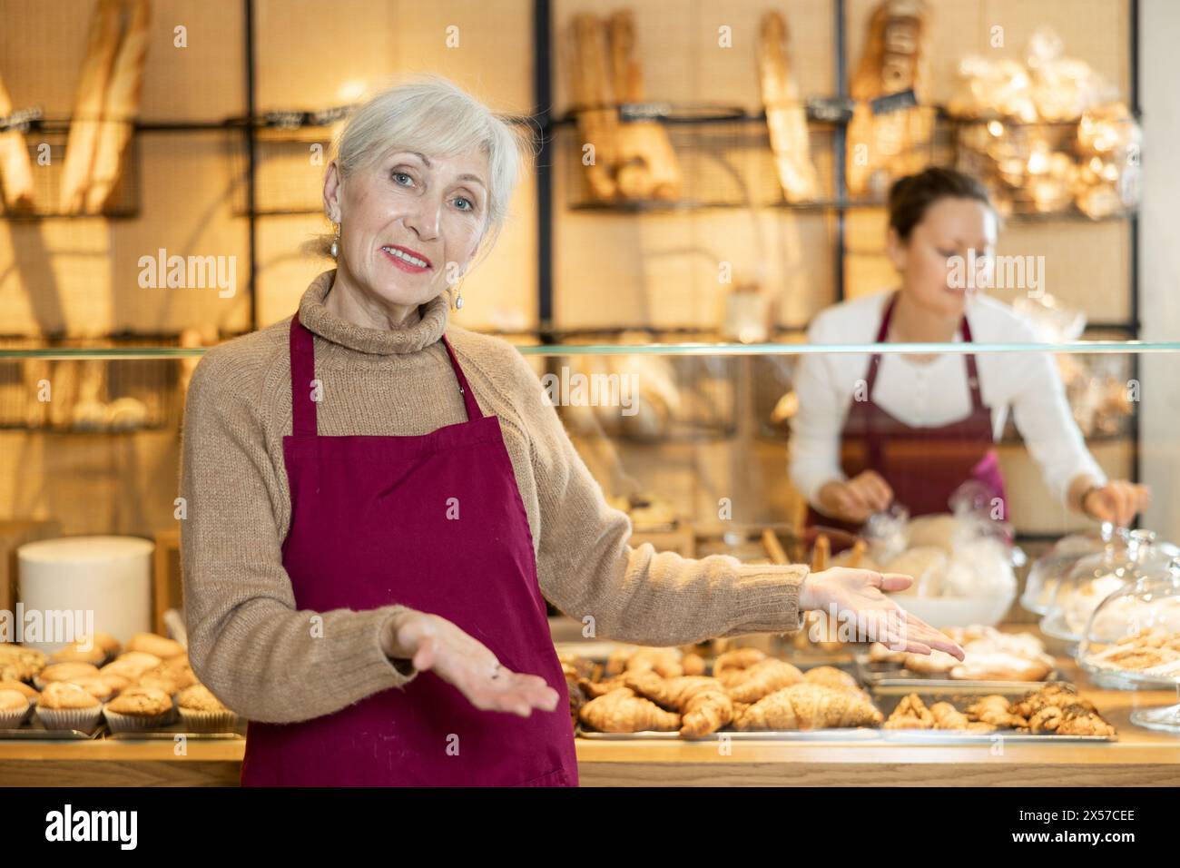 Elderly bakeshop saleswoman in maroon apron recommending baked goods Stock Photo - Alamy