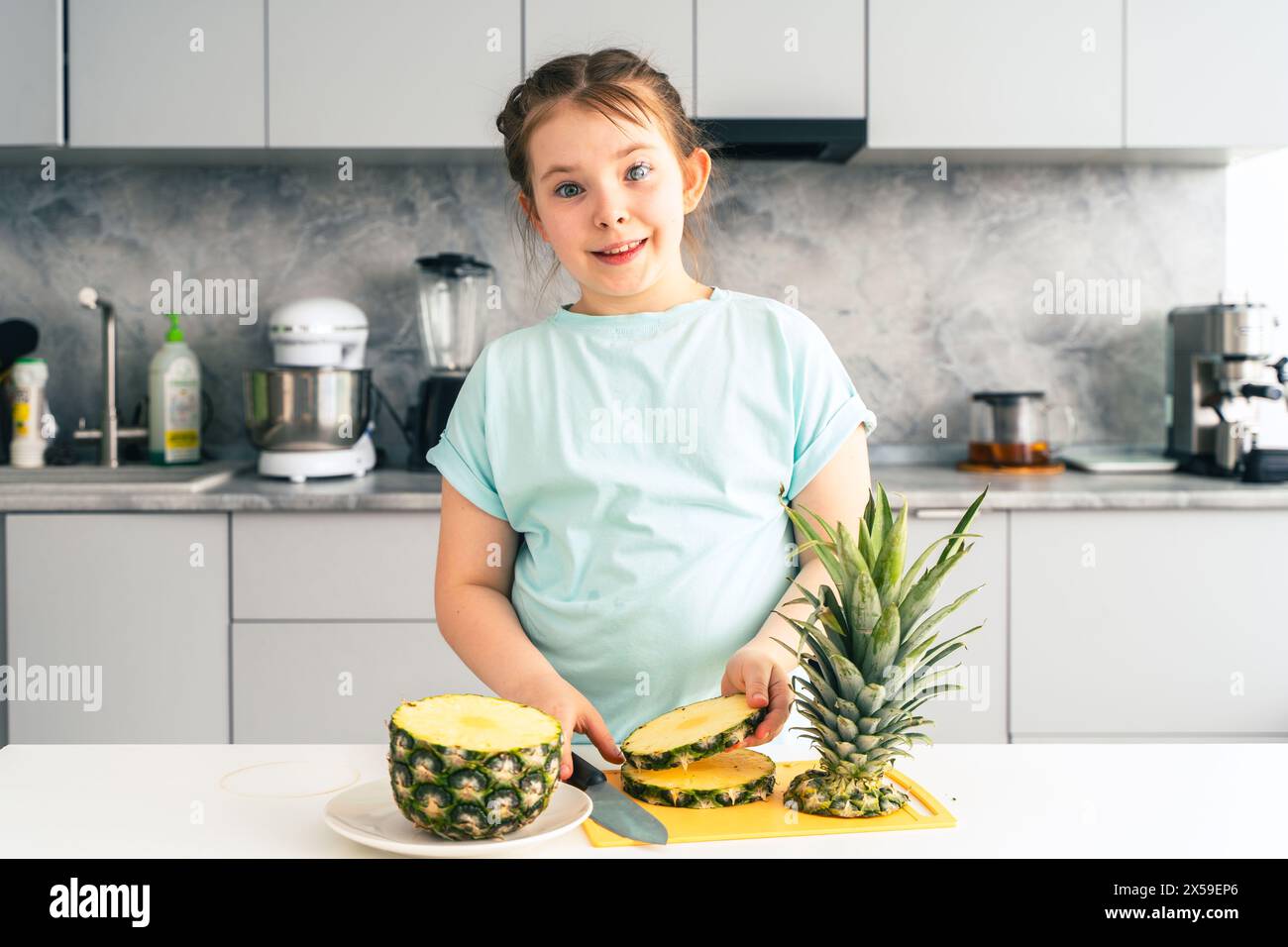 Happy little girl cutting pineapple on the background of the kitchen at home. Pre-teen girl ...