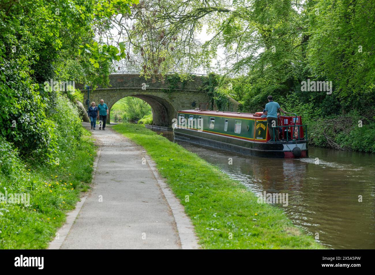 A hired narrowboat passes a couple walking along a canal towpath close ...