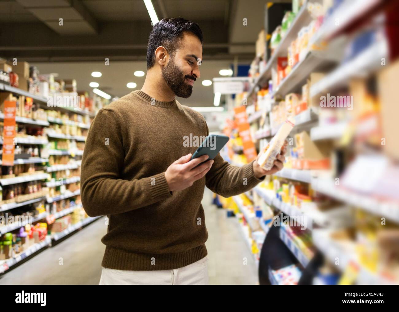 A smiling man browses grocery items while consulting his smartphone in ...