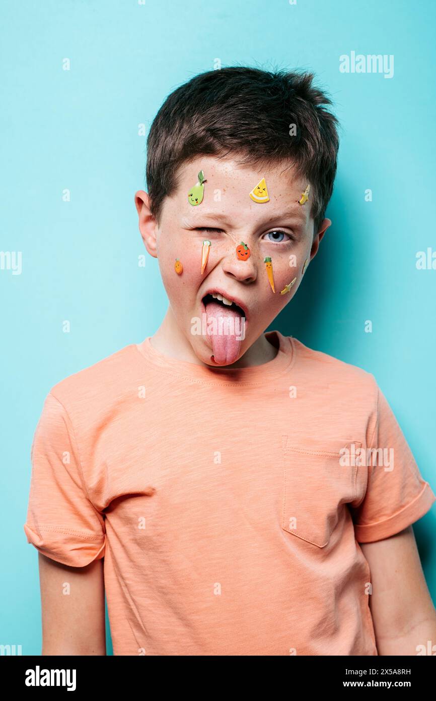 A young boy with quirky fruit-themed stickers playfully adorning his face sticks out his tongue ...