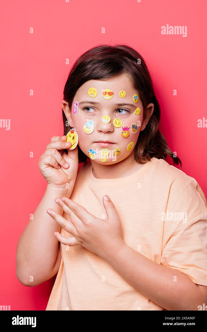 A young girl with emoji stickers on her face is holding one sticker ...