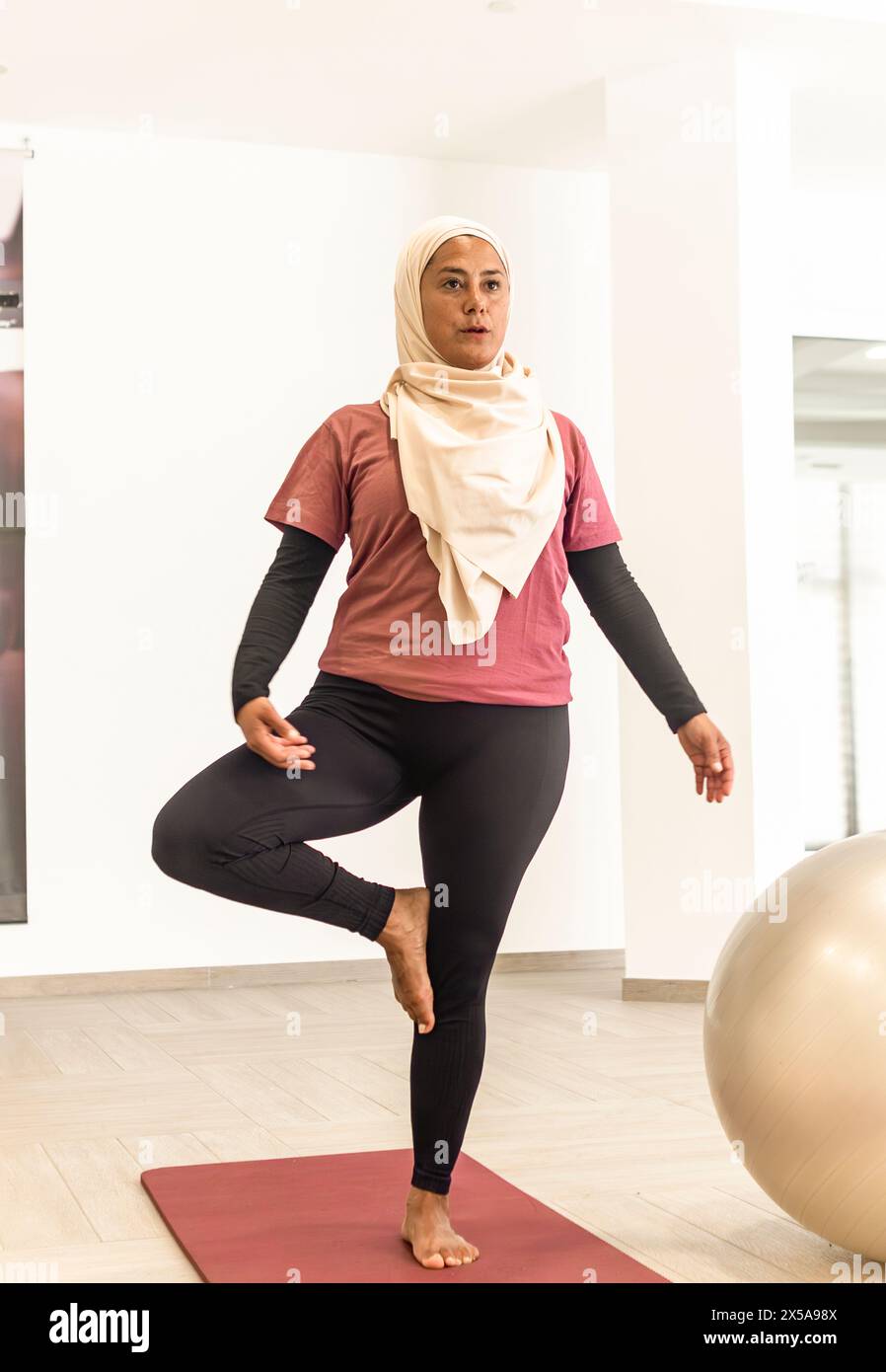 A focused Muslim woman in a hijab prepares for a yoga session by ...