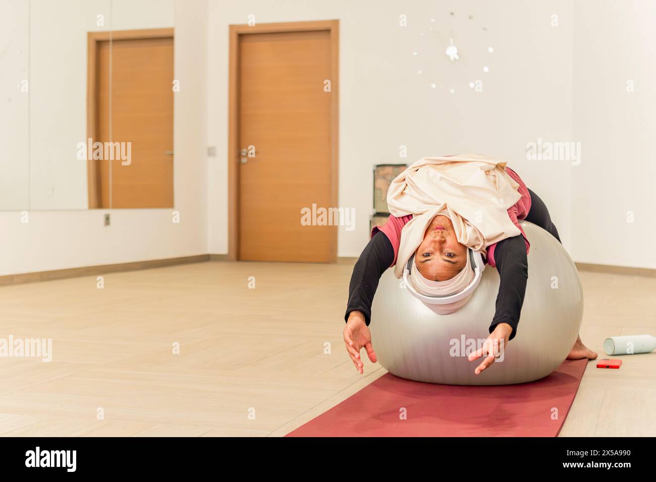A Muslim woman in a hijab engages in a yoga pose using a fitness ball ...
