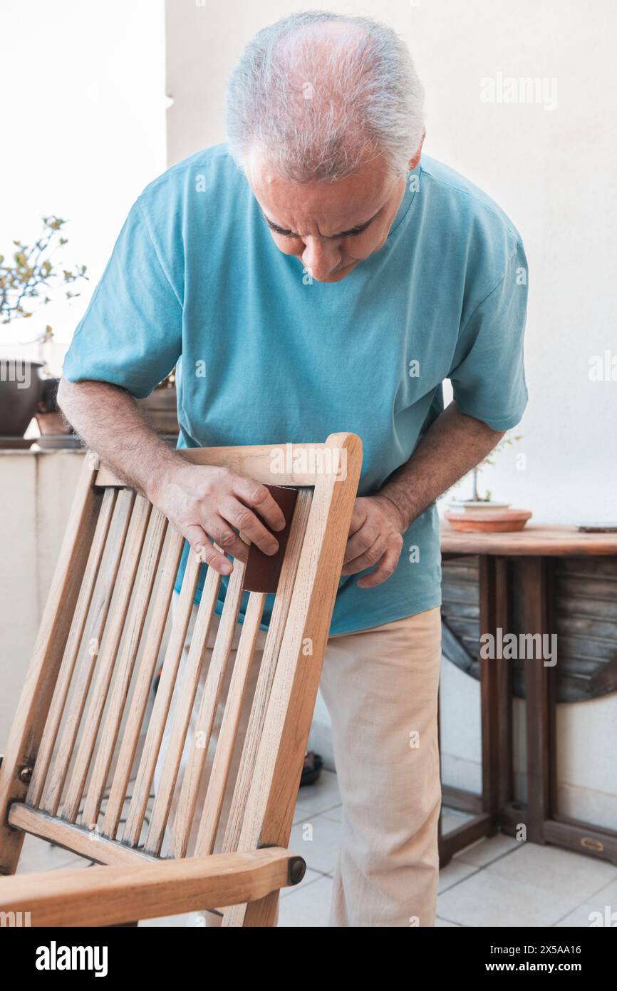 An elder craftsman sanding a wooden chair outdoors, focused on ...