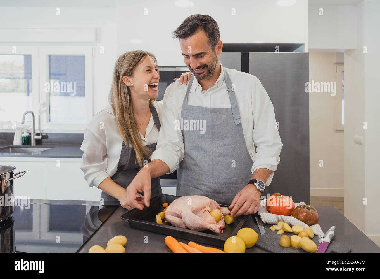 A happy couple shares a laugh while cooking a roast chicken with fresh ...