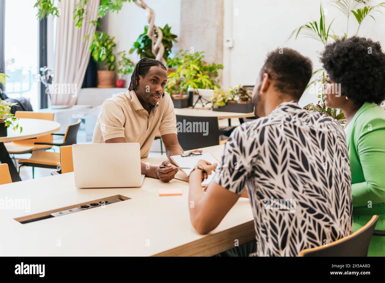 Three professionals engage in a collaborative discussion in a bright and plant-filled coworking ...