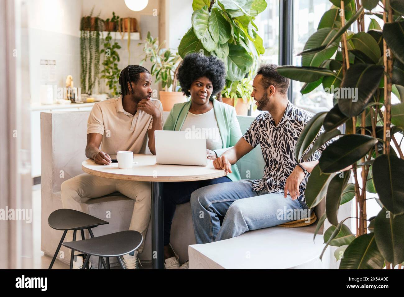 Three colleagues engage in discussion around a laptop in a bright and plant-filled coworking ...