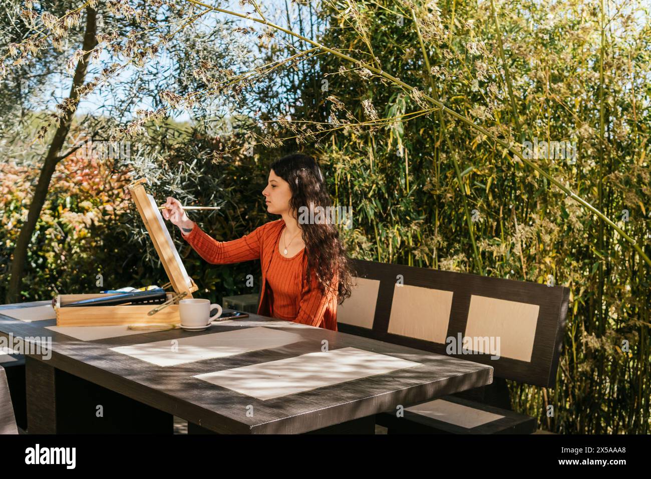 A focused woman artist paints on a canvas outdoors surrounded by lush greenery on a bright sunny ...