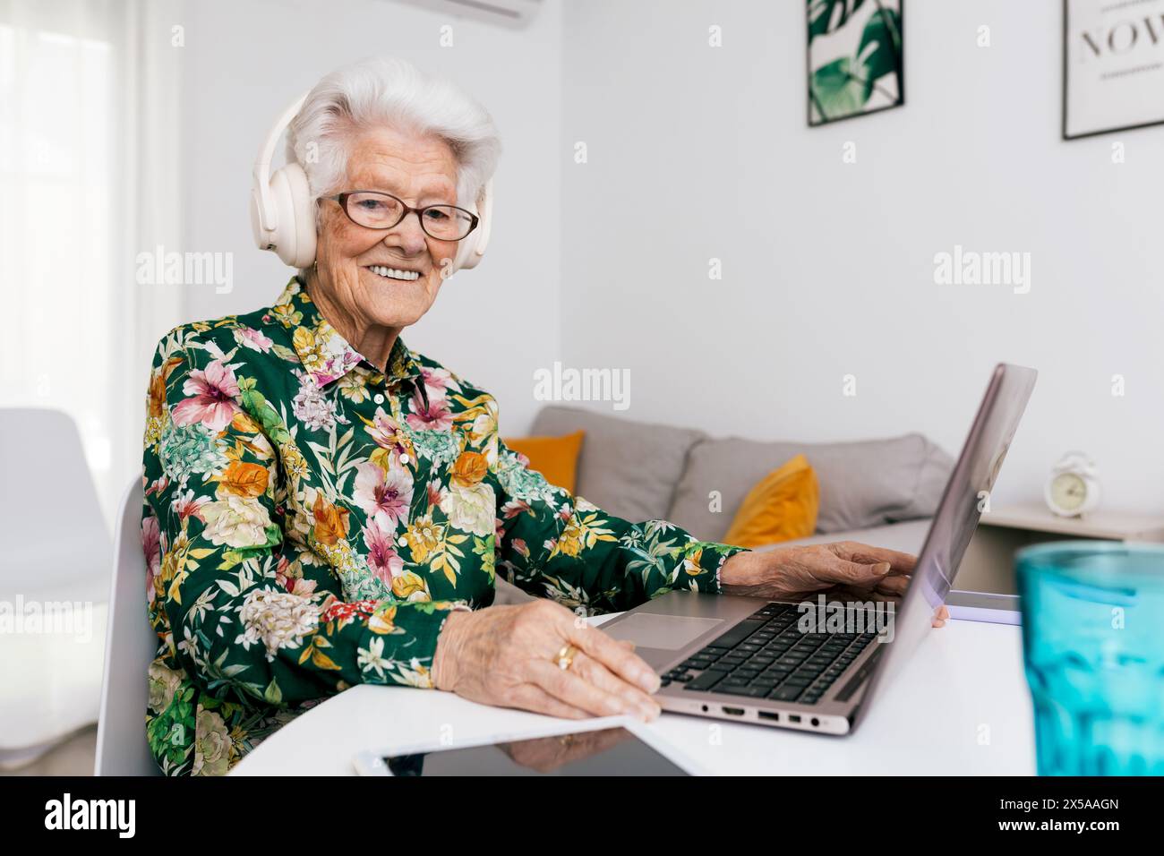 Joyful elderly lady with headphones uses a laptop in a bright, cozy ...