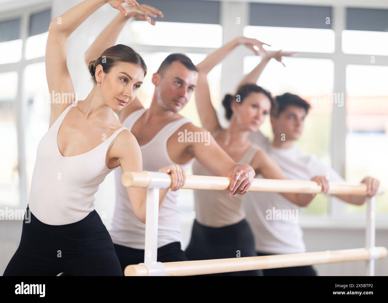 Diligent beautiful young female dancer exercising ballet moves at barre ...