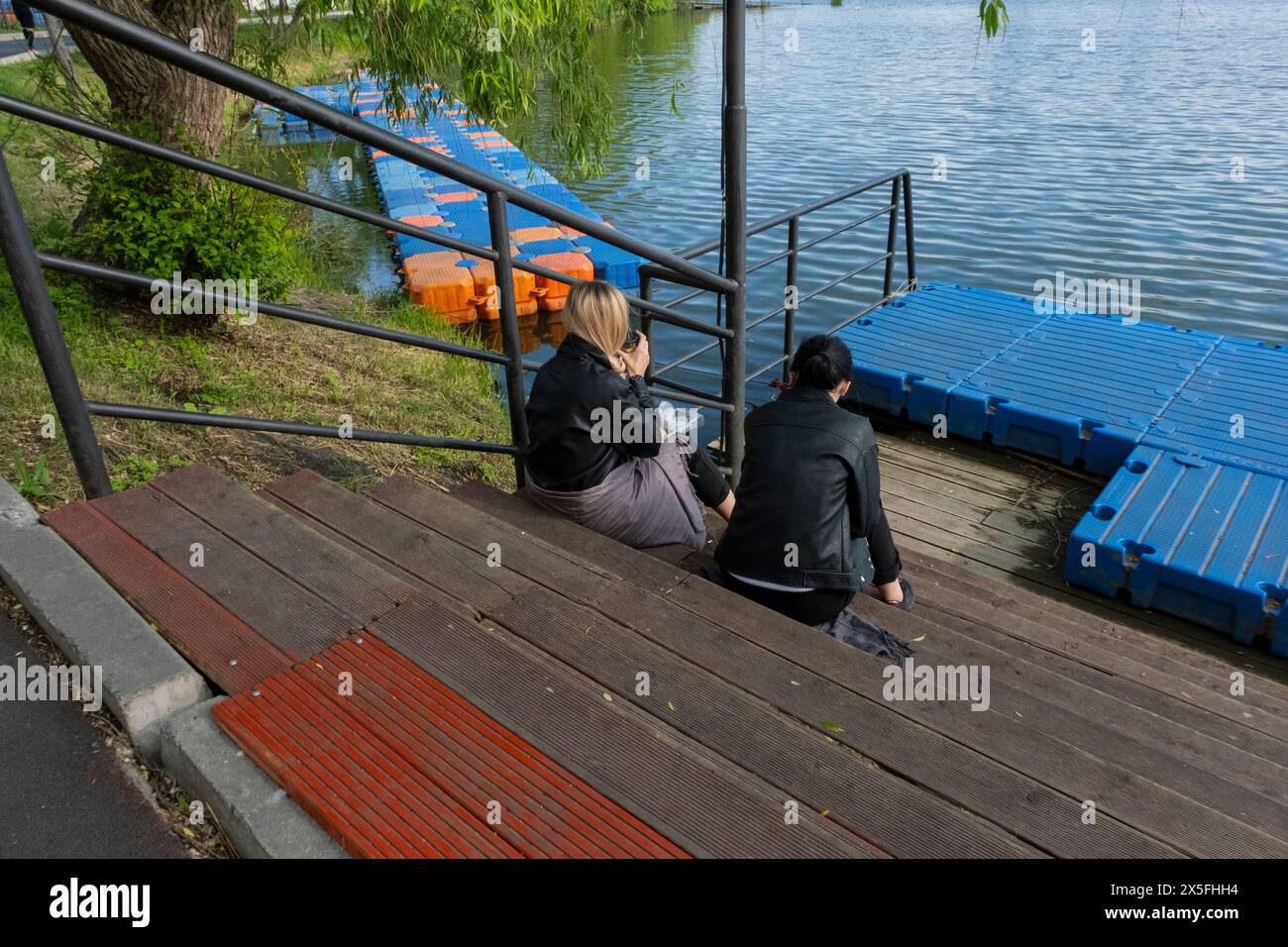 Two women sitting on the steps of a pier by a lake drinking coffee. Two ...