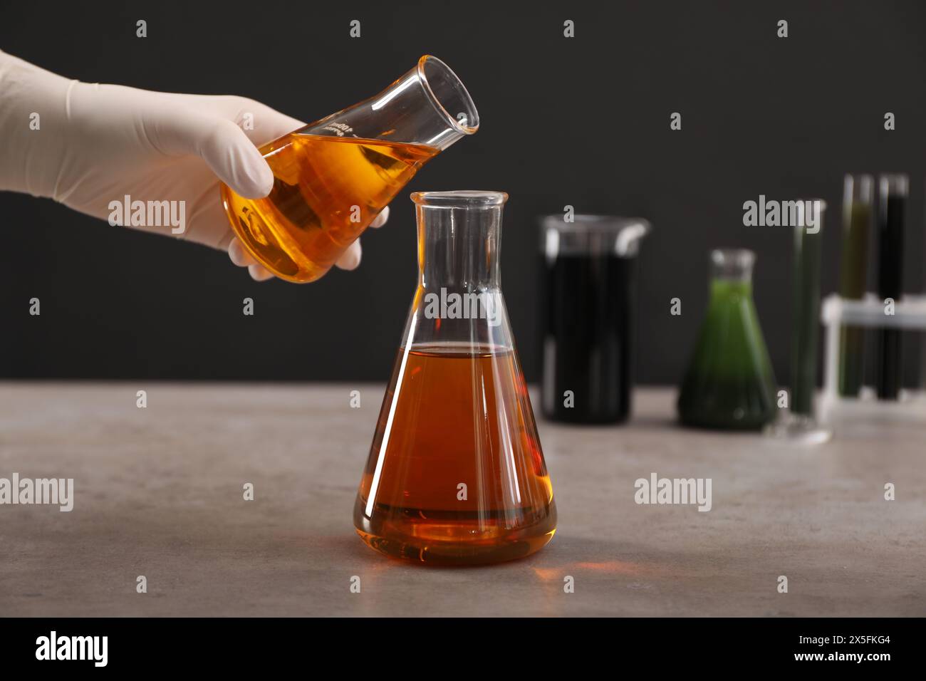 Woman pouring yellow crude oil into flask at grey table against dark ...