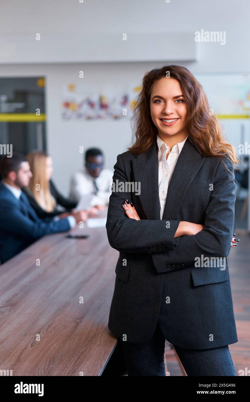 Confident blonde businesswoman stands in office, arms crossed, smiling at camera as diverse team ...