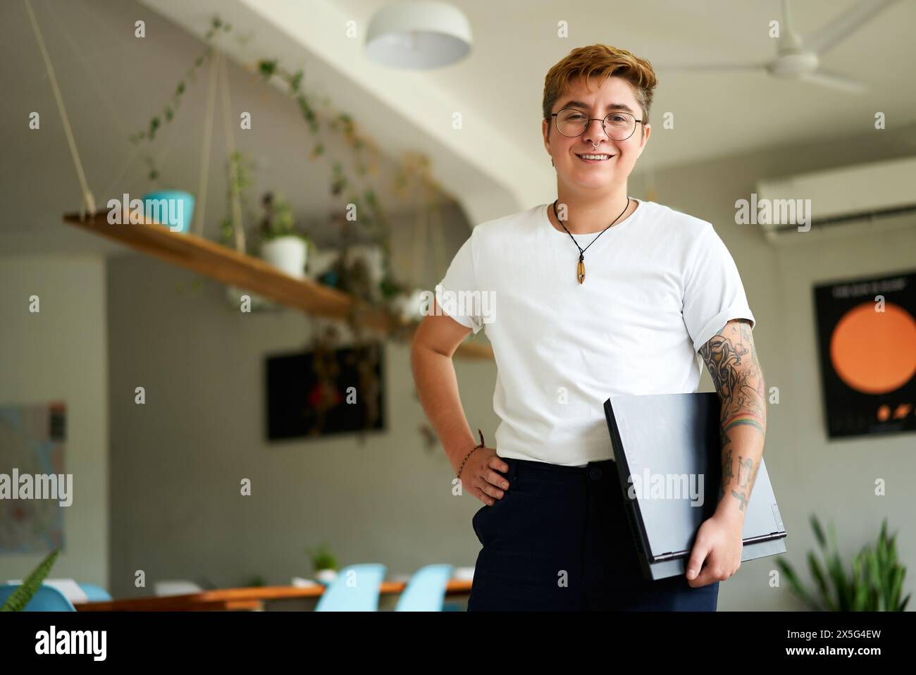 Confident transgender male employee stands with laptop in modern office ...